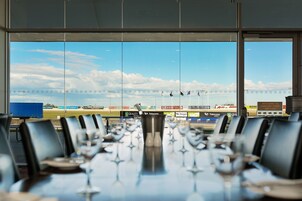 Sala de conferências com vista para um campo esportivo, mesas com cadeiras e uma janela grande.