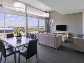 Quarto de hotel com área de refeições, sofá, televisão de tela plana e janelas amplas com vista para um campo esportivo.