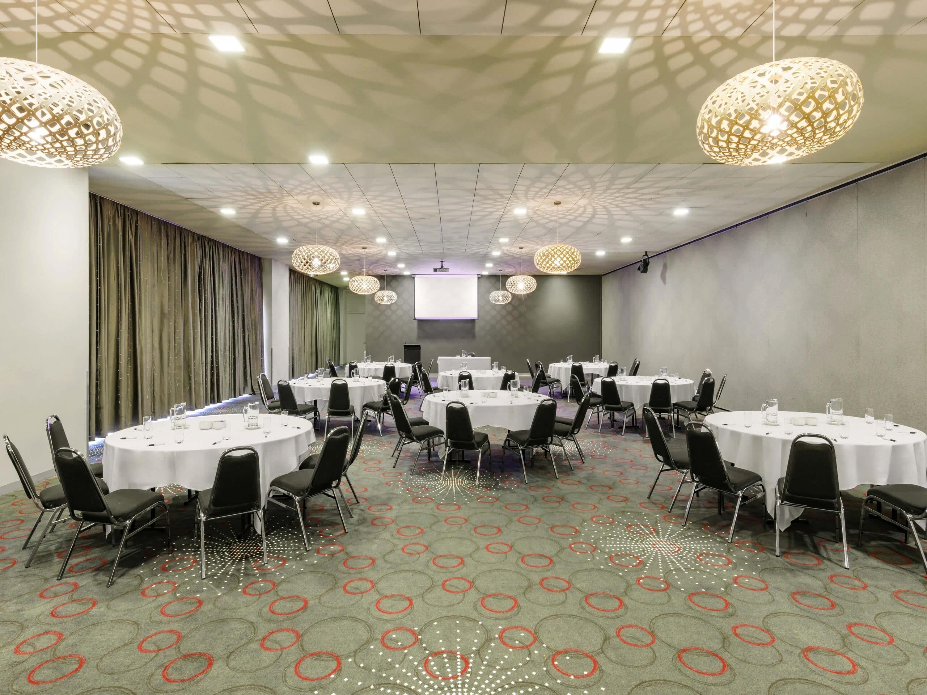 Sala de conferências com mesas redondas, cadeiras pretas e carpete estampado.