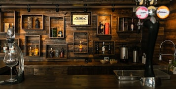 Uma área de bar com prateleiras de madeira exibindo diversas garrafas de bebidas alcoólicas, um letreiro para BBC Cerveza Fresca de Barril e uma cafeteira.