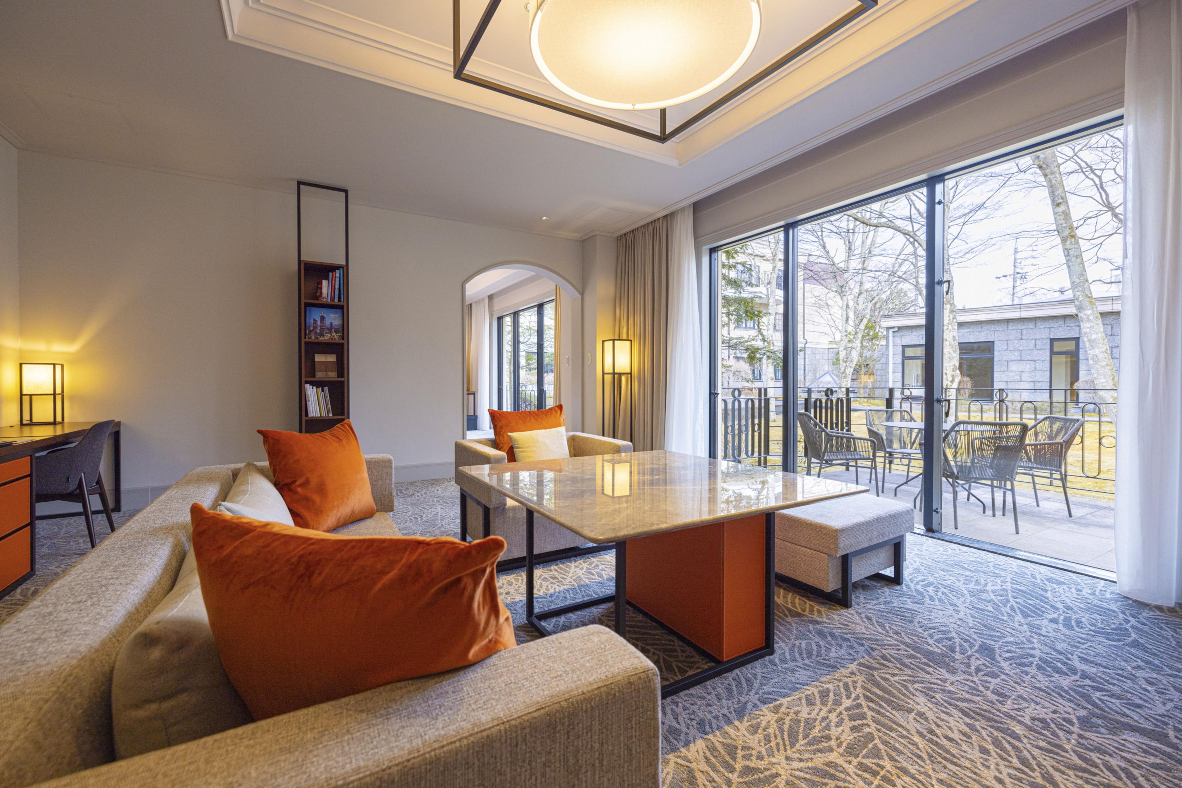 Quarto de hotel com sofá, mesa de jantar, estante de livros e uma grande janela com vista para o exterior.