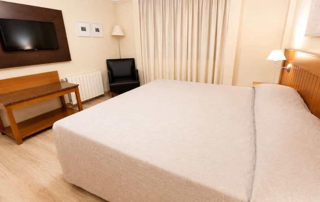 Quarto de hotel com cama, televisão presa na parede, escrivaninha e uma cadeira.