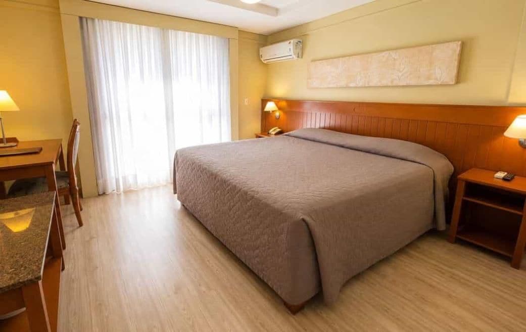 Quarto de hotel com uma cama grande, uma escrivaninha com cadeira, mesa de cabeceira com abajur e janela com cortinas.