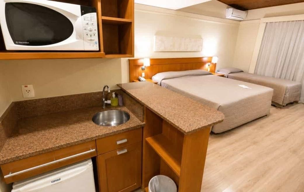 Cozinha compacta equipada com micro-ondas, pia e armários. Há uma cama com encosto e mesa de cabeceira com luminária.