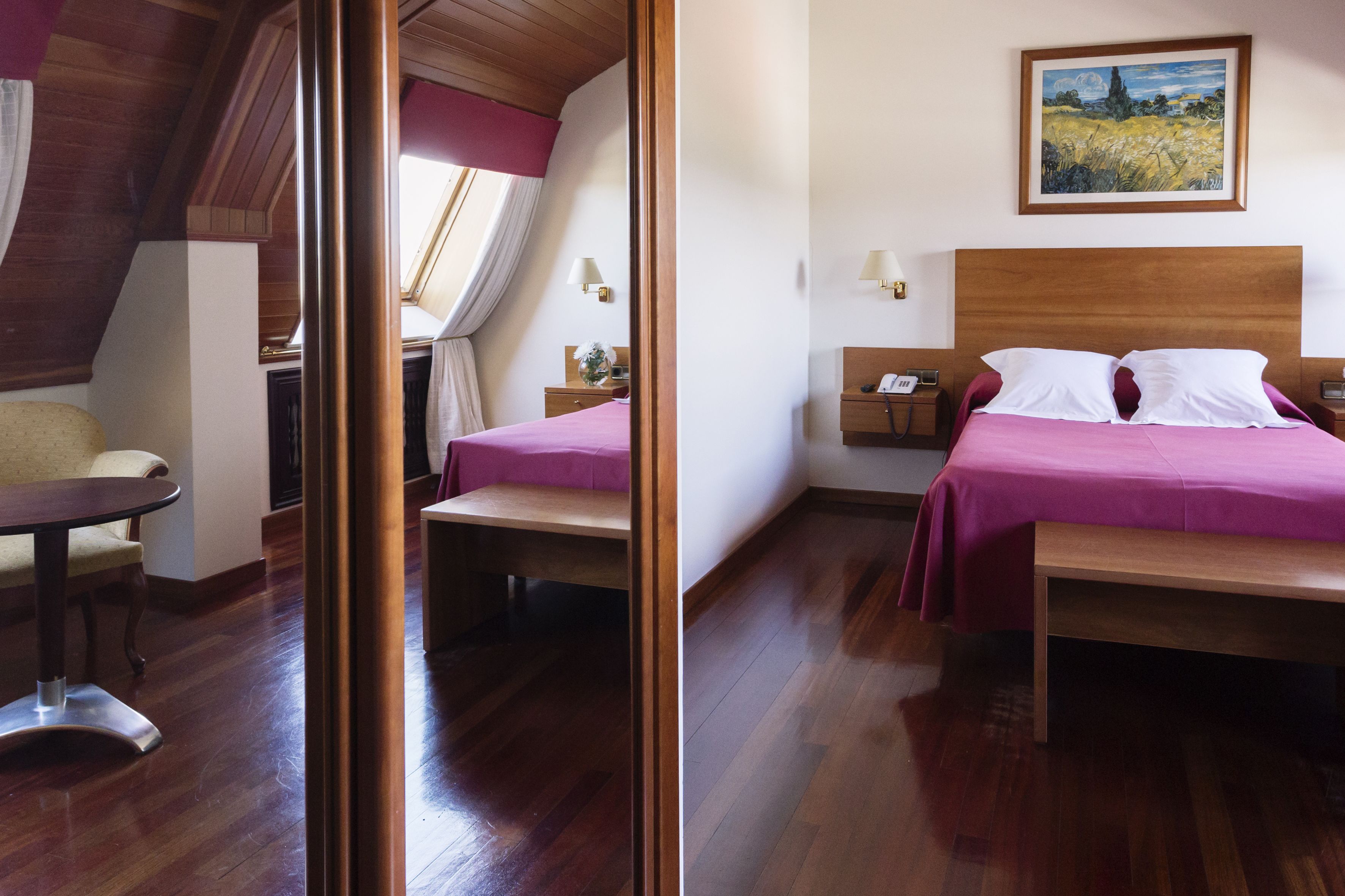 Quarto de hotel com teto de madeira, cama com roupa de cama rosa, um banco de madeira, uma mesinha e um quadro na parede.