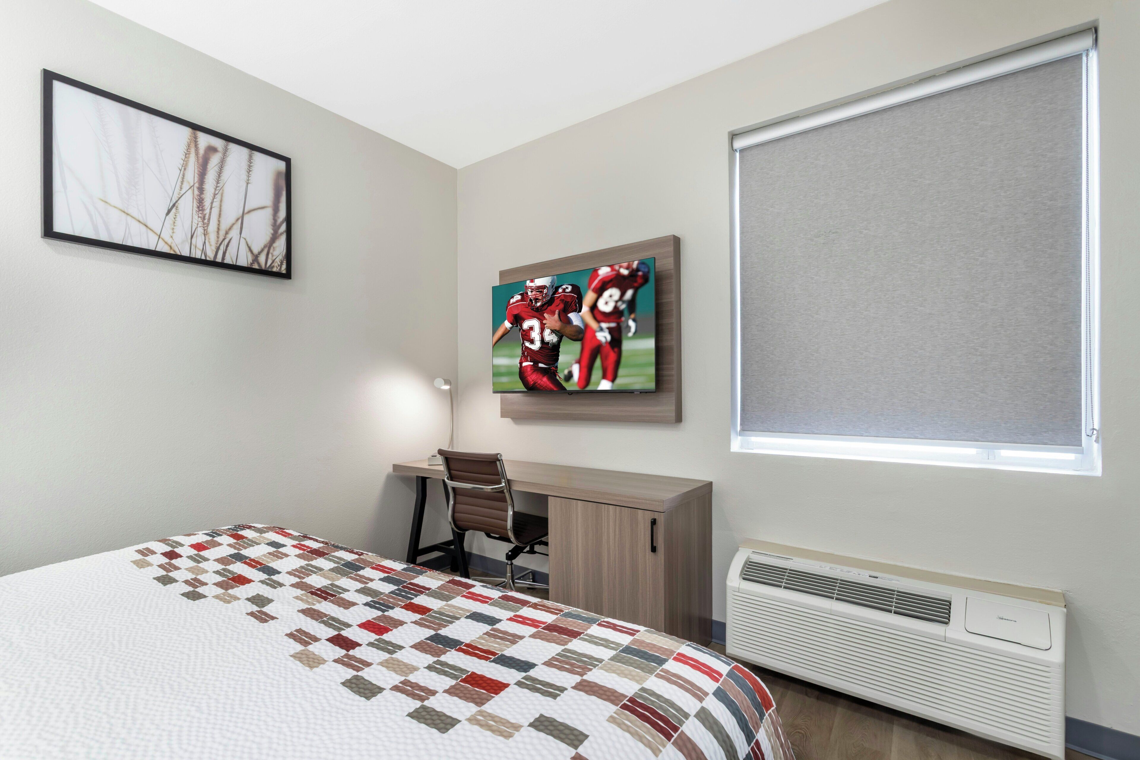 Quarto com cama, escrivaninha, cadeira, pintura, janela e ar-condicionado.