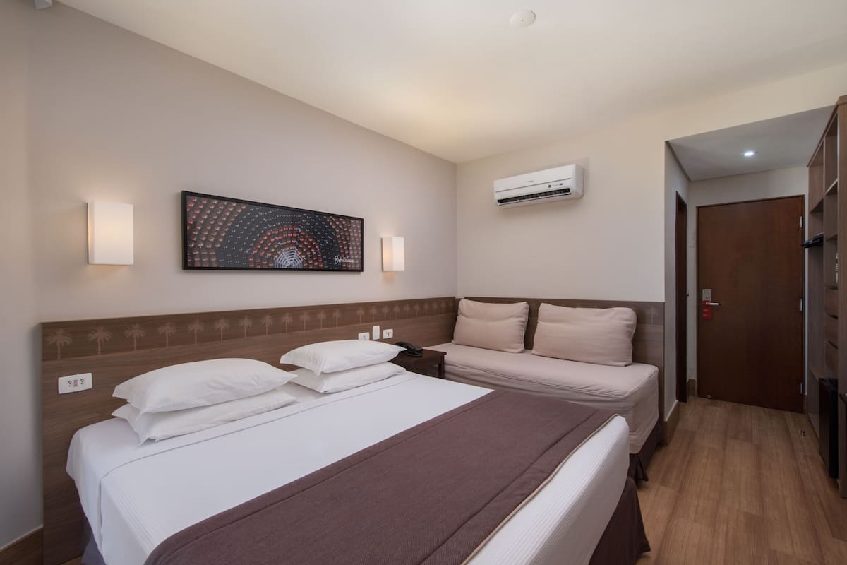 Quarto de hotel com uma cama grande, uma cama menor, uma mesa de cabeceira, um ar condicionado fixado na parede e um quadro na parede.