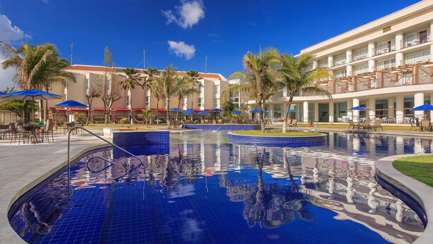 Uma área de piscina de hotel com vários níveis, palmeiras e área de estar ao ar livre sob guarda-sóis.