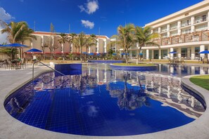 Uma área de piscina de hotel com vários níveis, palmeiras e área de estar ao ar livre sob guarda-sóis.