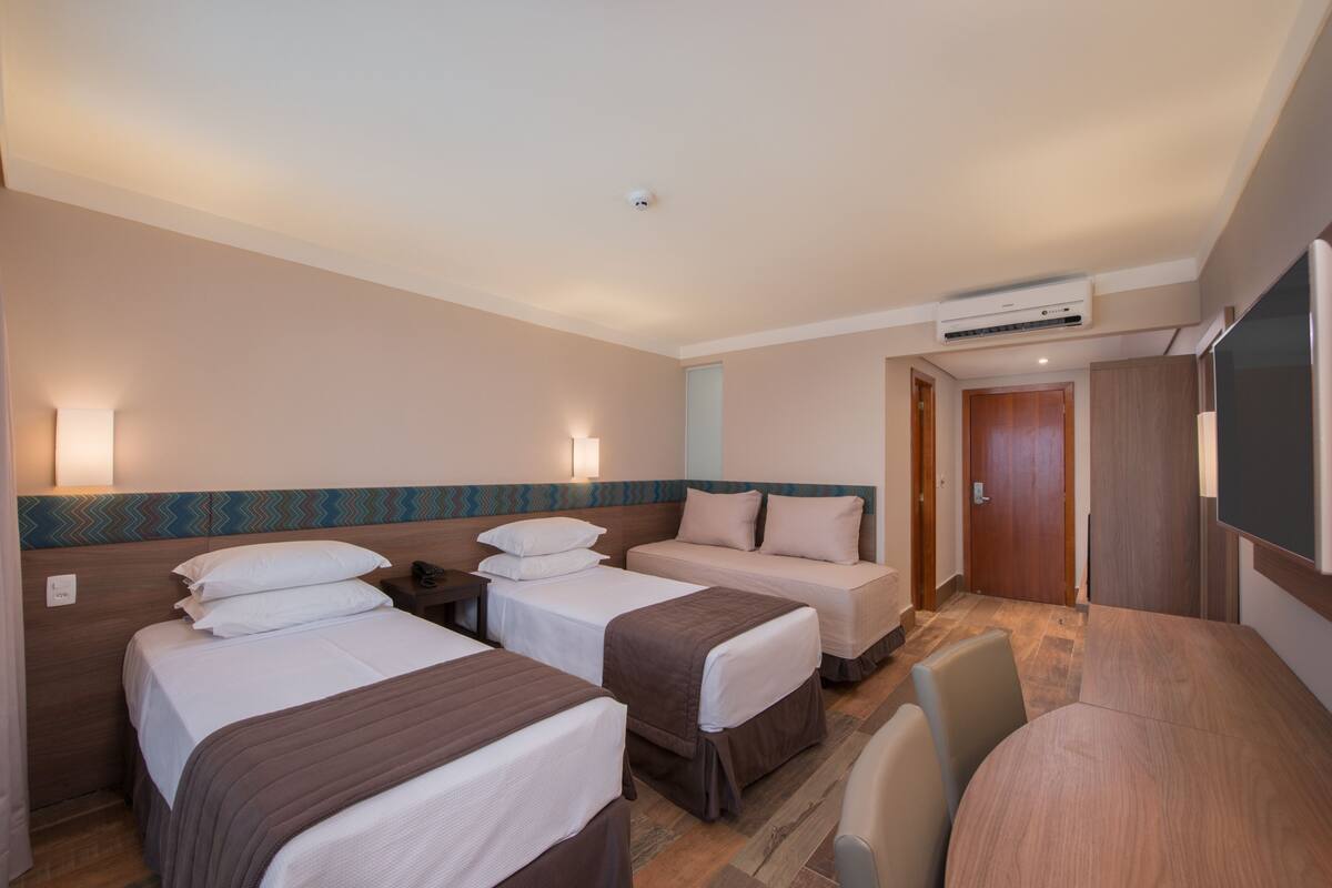 Quarto de hotel com duas camas, uma escrivaninha, uma cadeira, uma televisão e ar condicionado.