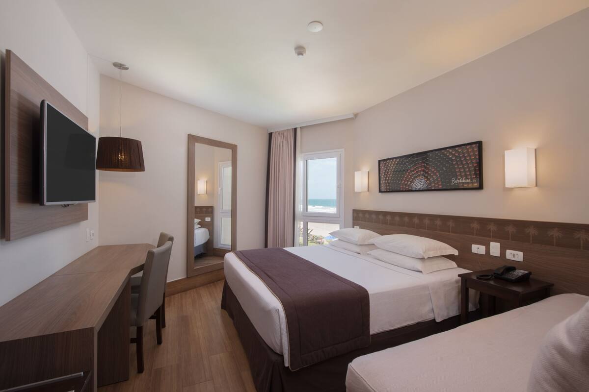 Quarto de hotel com cama, escrivaninha, cadeira, televisão, mesa de cabeceira com telefone e janela com vista.