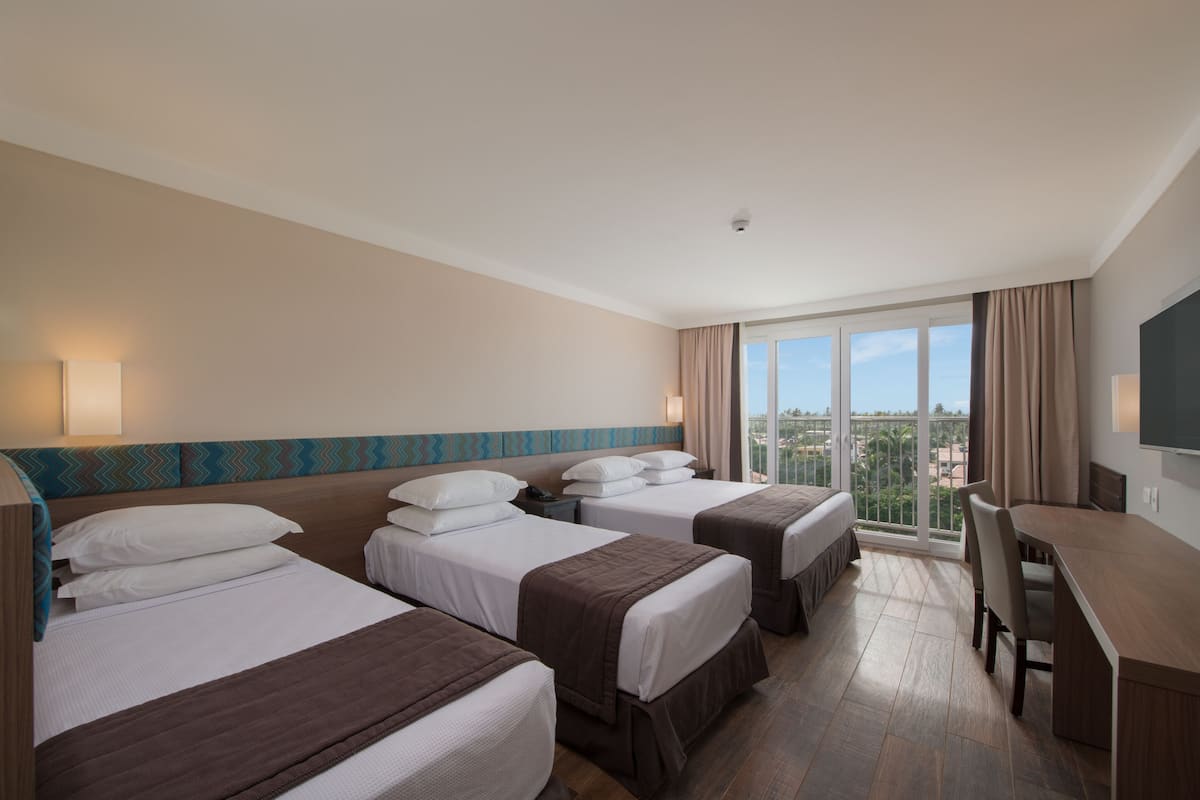 Quarto de hotel com três camas, uma escrivaninha e uma janela grande com vista para a cidade.