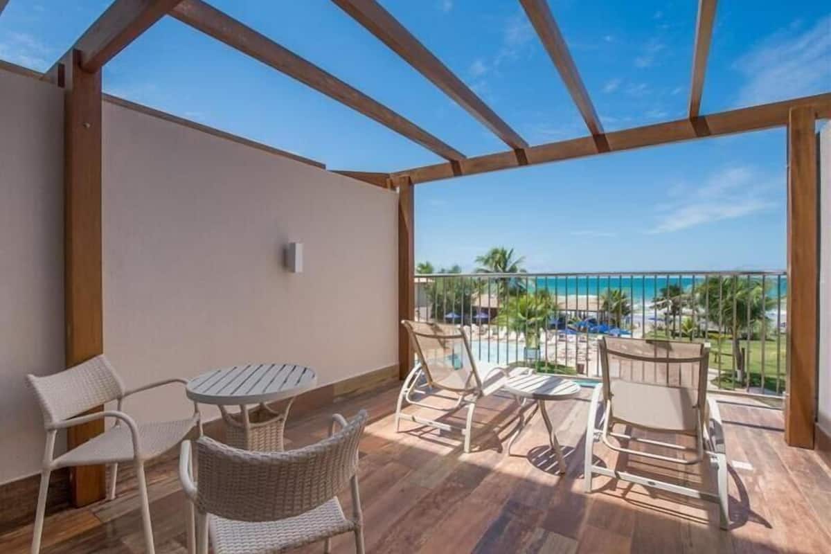 Área externa coberta com mesa e cadeiras, vista para uma praia e palmeiras, e estrutura de teto de madeira.