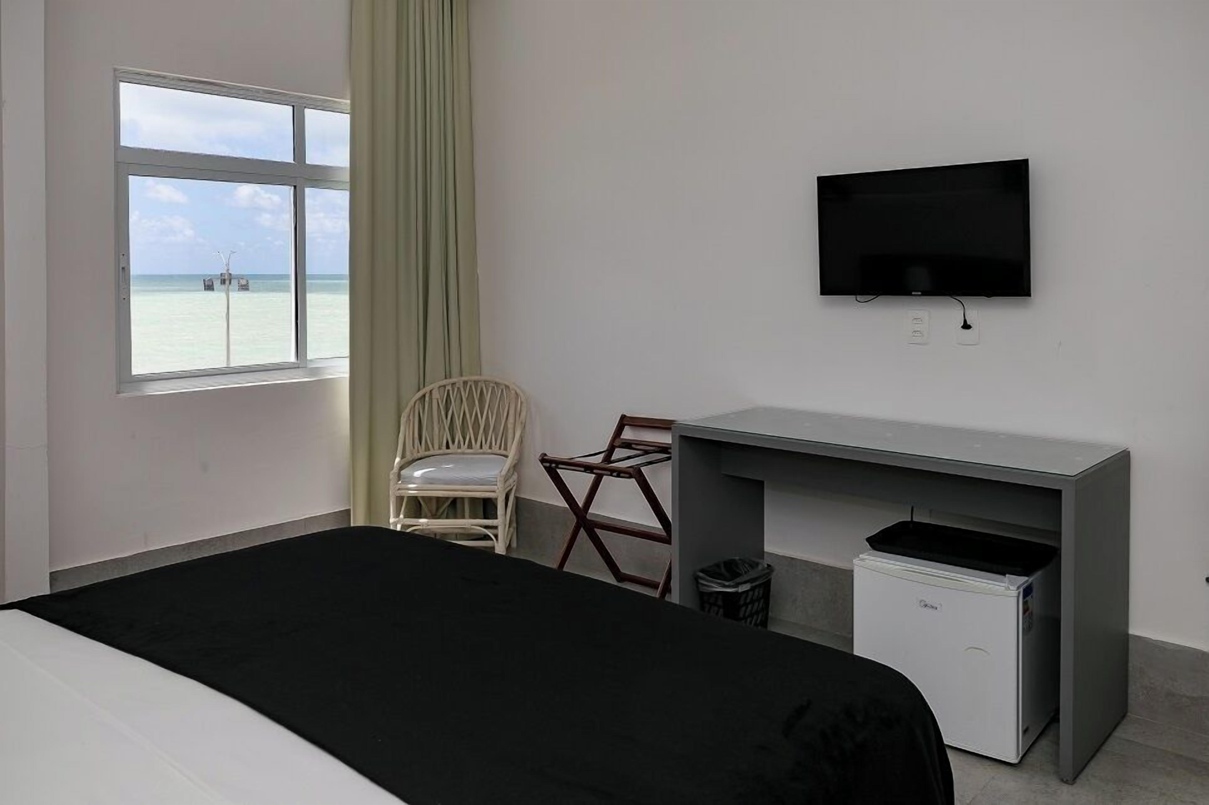 Quarto de hotel com cama, escrivaninha, cadeira, televisão e vista para o mar.
