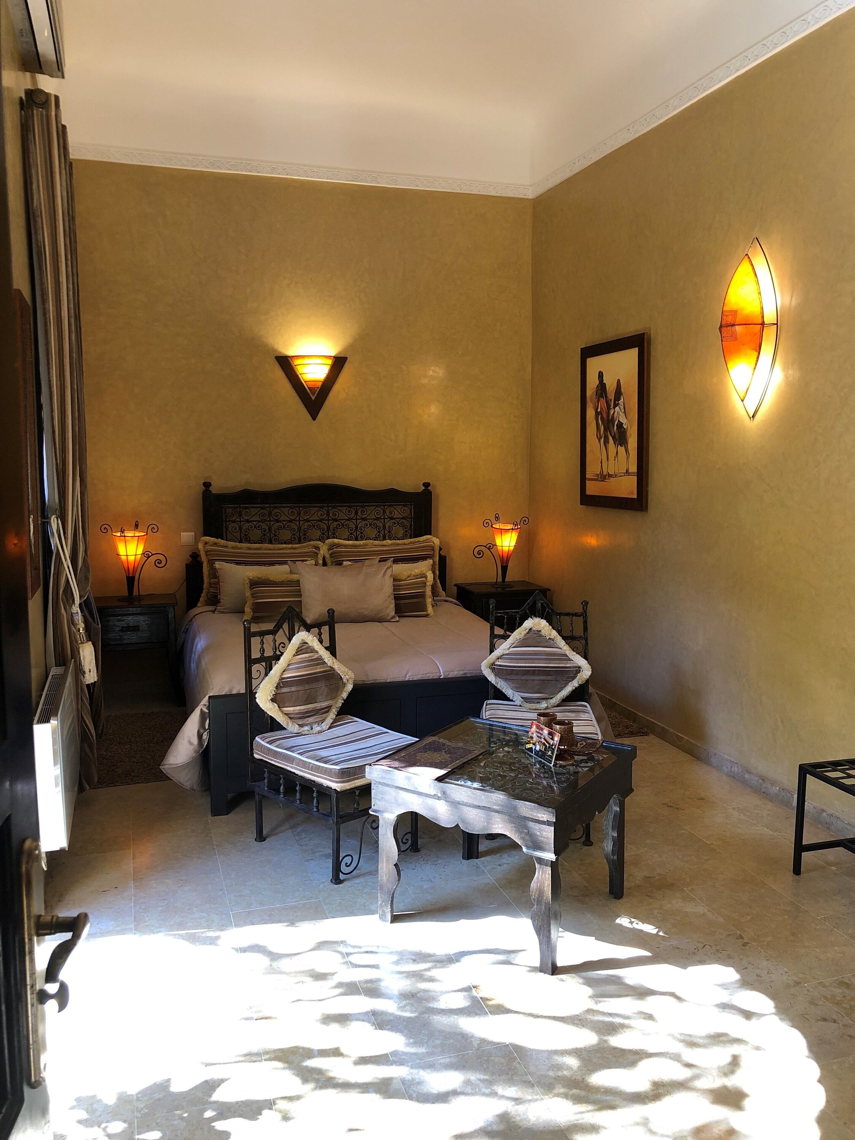 Quarto de hotel com cama, duas mesas de cabeceira, uma mesinha com abajur, um quadro na parede e vista para o exterior.