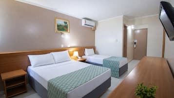 Quarto de hotel com duas camas, uma escrivaninha de madeira, televisão, uma pequena planta e um aparelho de ar condicionado.