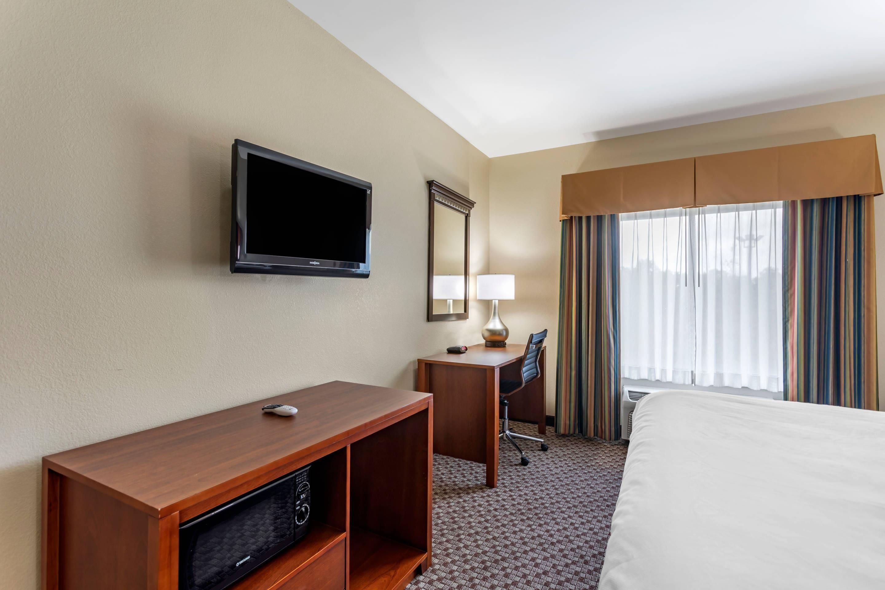 Quarto de hotel com cama, escrivaninha, televisão, espelho, abajur e janela com cortinas.