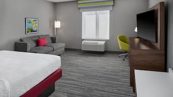Quarto de hotel com cama, sofá, poltrona, televisão, janela com persianas e radiador.