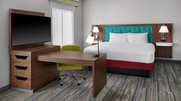 Quarto de hotel com escrivaninha de madeira, televisão de tela plana, cama com cabeceira verde-água, duas luminárias de cabeceira e uma cadeira verde.