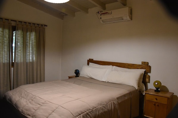 Quarto com cama de madeira, roupa de cama branca, mesa de cabeceira com um globo e um aparelho de ar condicionado na parede.