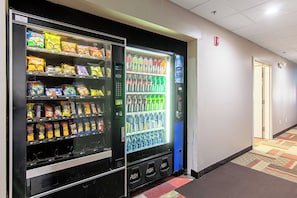 Máquina de vendas automática com lanches e bebidas, situada em um corredor com piso em carpete e uma porta ao fundo.
