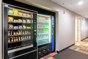 Máquina de vendas automática com lanches e bebidas, situada em um corredor com piso em carpete e uma porta ao fundo.