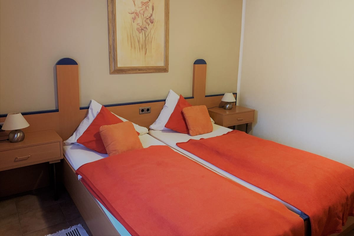 Uma cama de casal com roupa de cama laranja, duas mesas de cabeceira com luminárias e uma obra de arte emoldurada na parede.