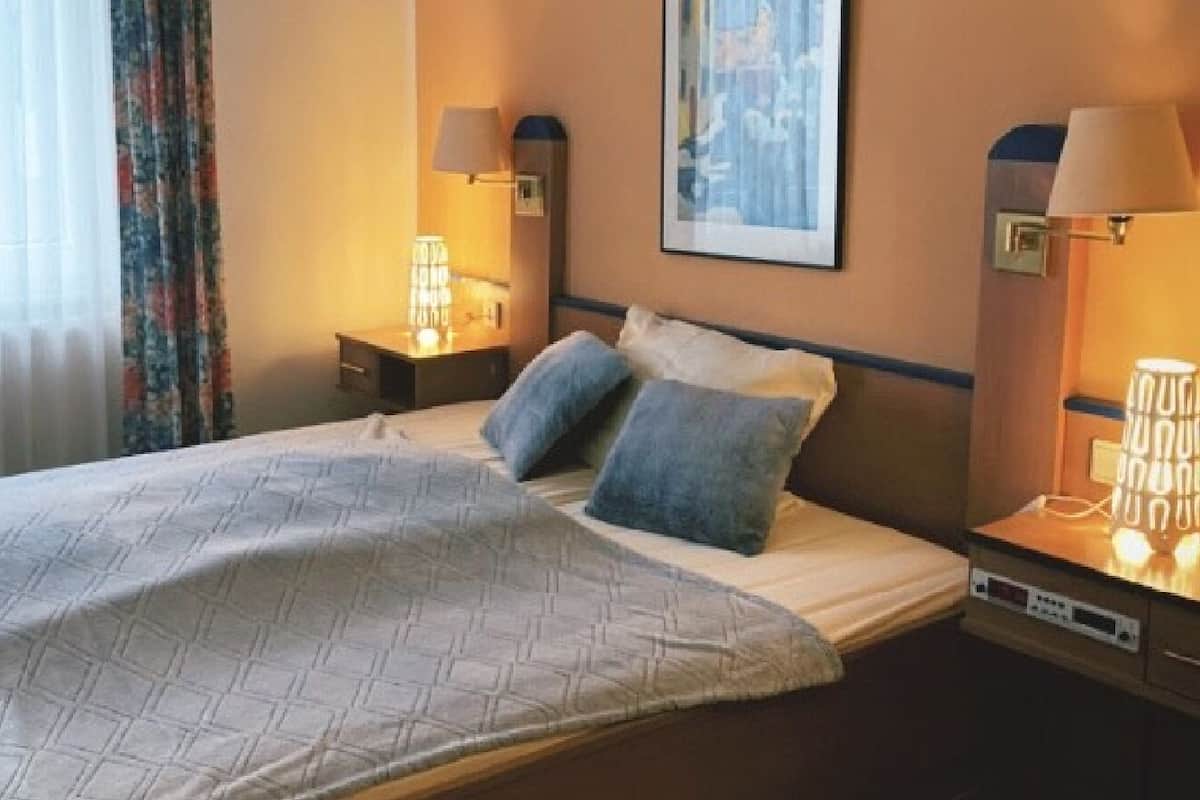 Quarto de hotel com uma cama, duas mesas de cabeceira com luminárias, uma janela com cortinas e um quadro na parede.