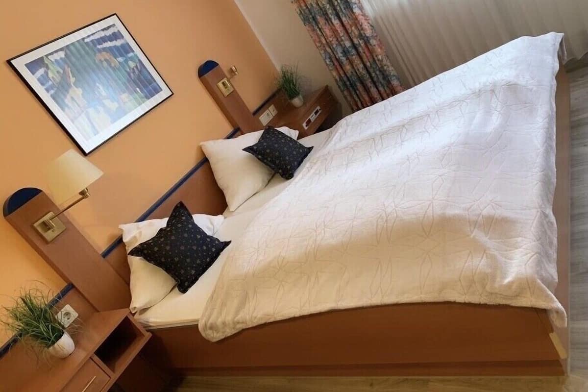 Cama bem arrumada com edredom branco e duas almofadas escuras, mesa de cabeceira com abajur e um quadro na parede.