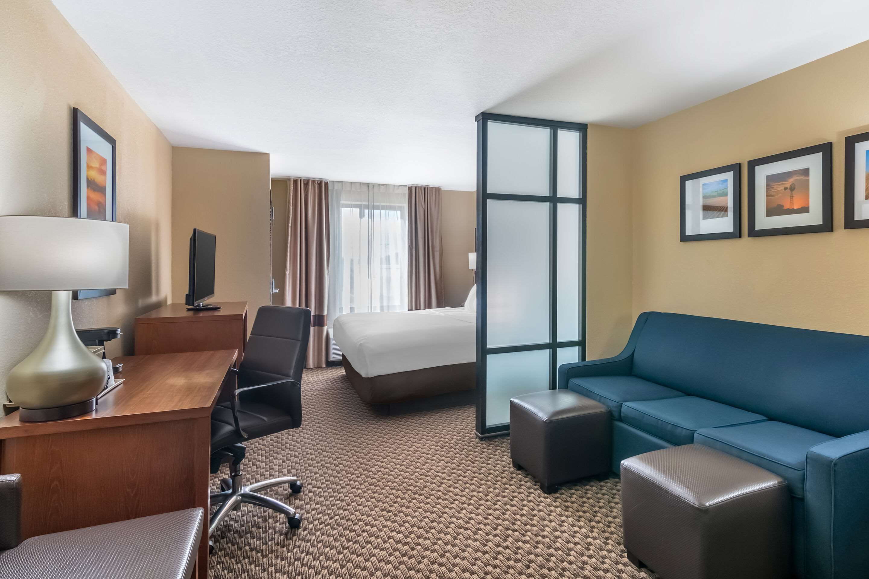 Quarto de hotel com cama, escrivaninha com luminária, sofá azul e televisão.
