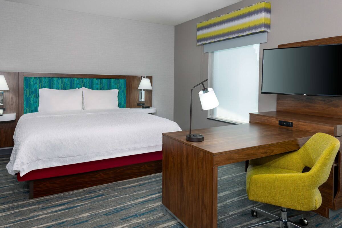 Quarto de hotel com cama, escrivaninha, cadeira, televisão e janela.
