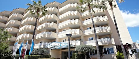 Um hotel com vários andares, varandas, palmeiras e portão fechado.