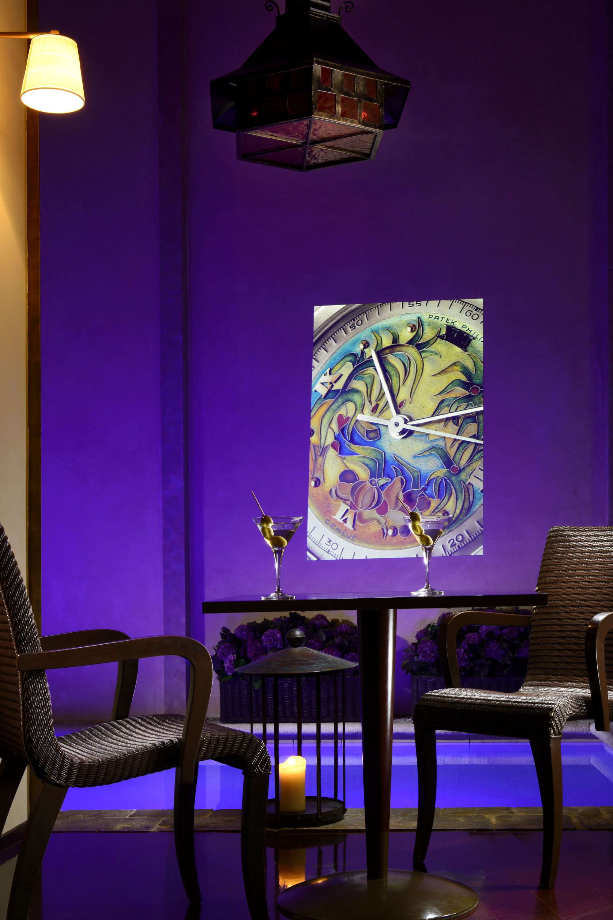 Quarto com paredes roxas, uma mesa com duas cadeiras, um relógio e uma vela.