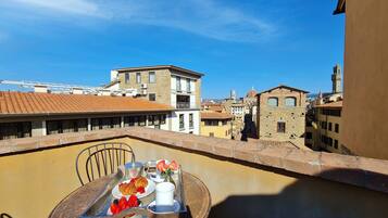 Varanda com mesa posta para café da manhã, vista para paisagem urbana com edifícios históricos e céu azul claro.