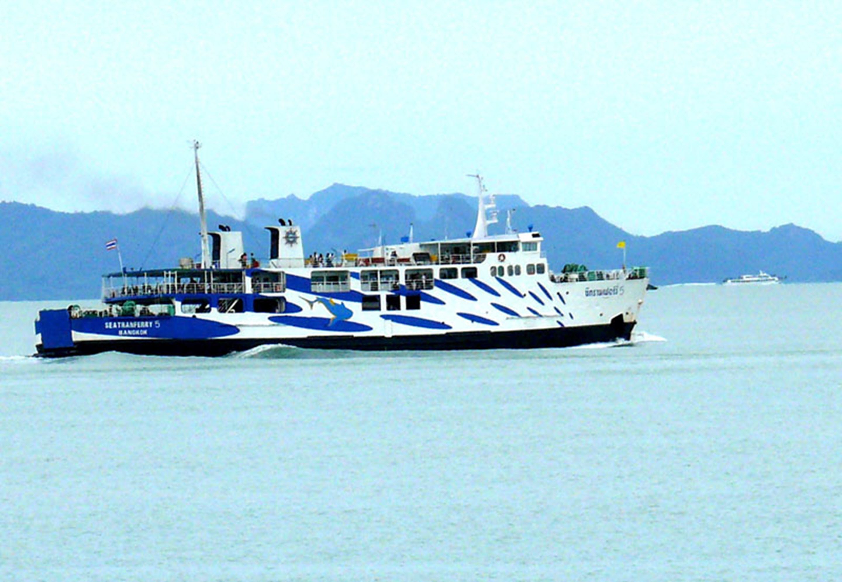 Um ferry com esquema de cores azul e branco, estrutura de cabines e vista para a água e montanhas ao fundo.