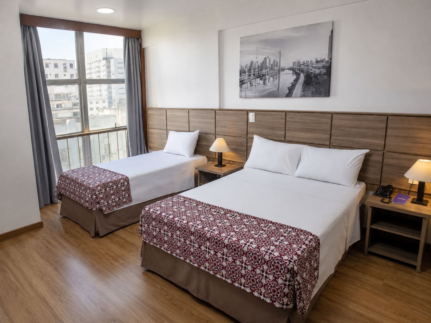 Quarto de hotel com duas camas, vista para a cidade, uma moldura na parede e janela com cortinas.