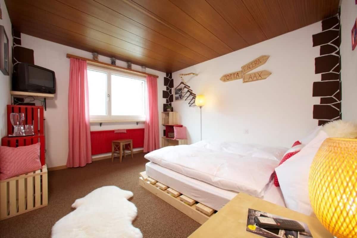 Um quarto aconchegante com teto de madeira, uma cama com roupa de cama branca, uma televisão em uma prateleira, uma janela com cortinas rosa e uma parede com placas de sinalização.