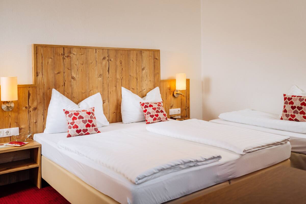 Quarto de hotel com cabeceira de madeira, duas camas, roupa de cama branca e almofadas com padrão de coração.