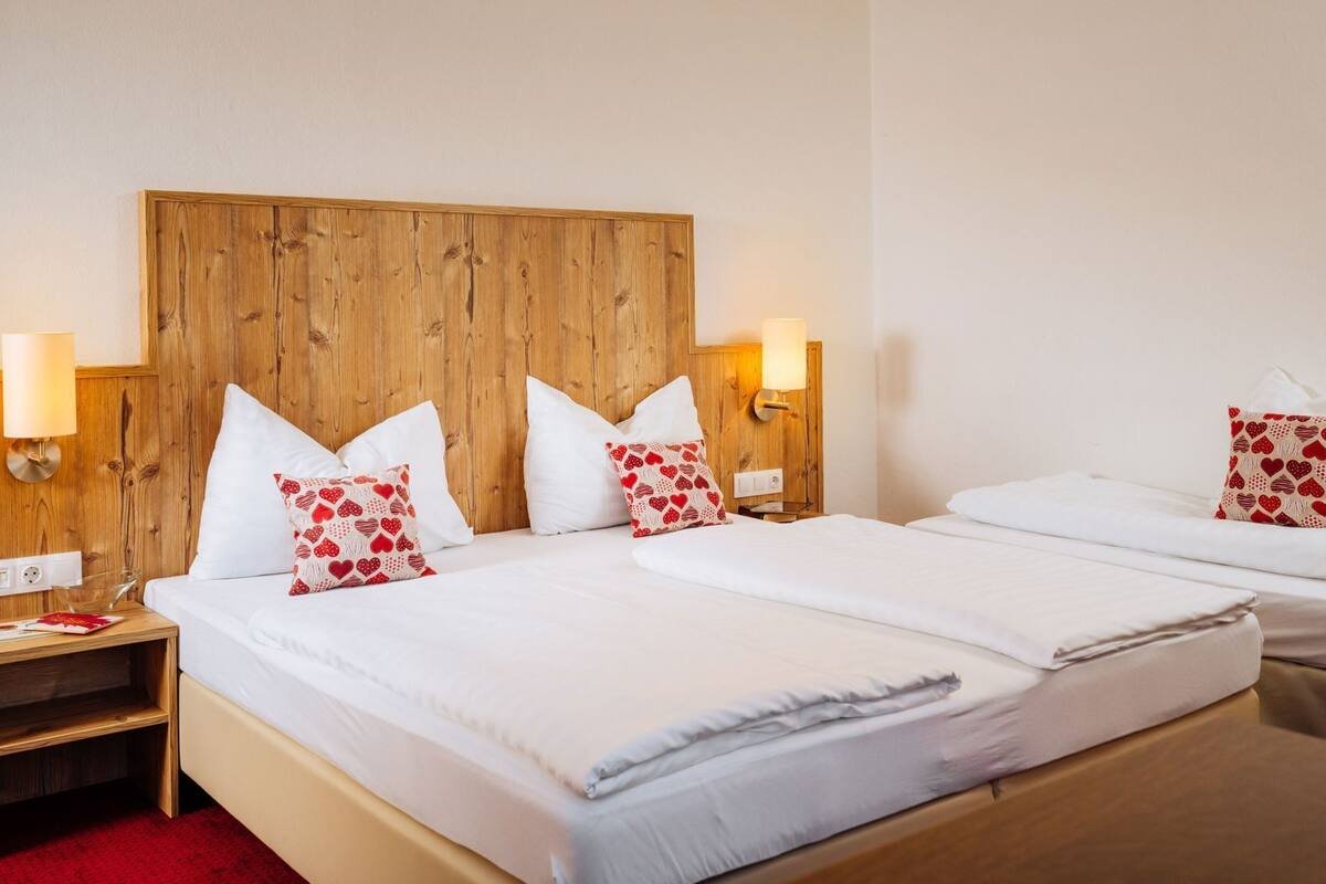 Quarto de hotel com cabeceira de madeira, duas camas, roupa de cama branca e almofadas com padrão de coração.