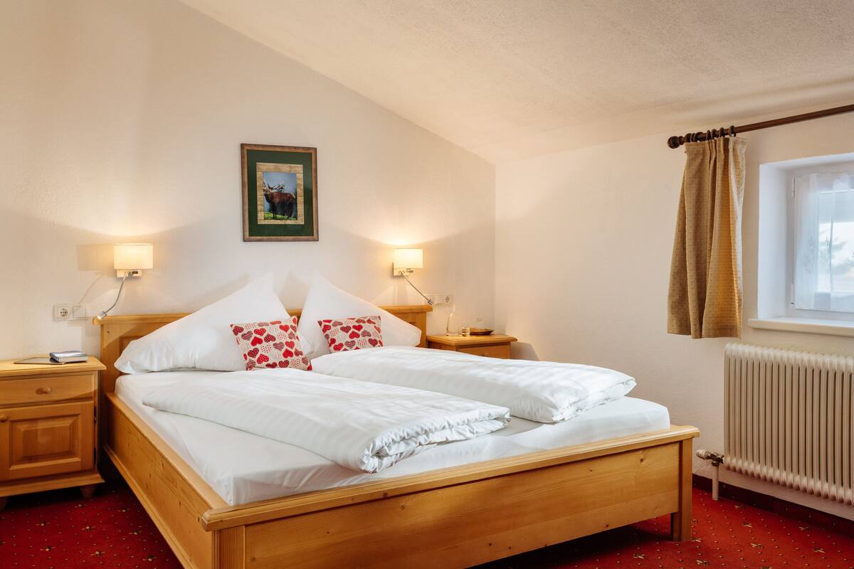 Uma cama de casal com roupa de cama branca, duas mesas de cabeceira com abajures, um quadro na parede, uma janela com cortina e um radiador.