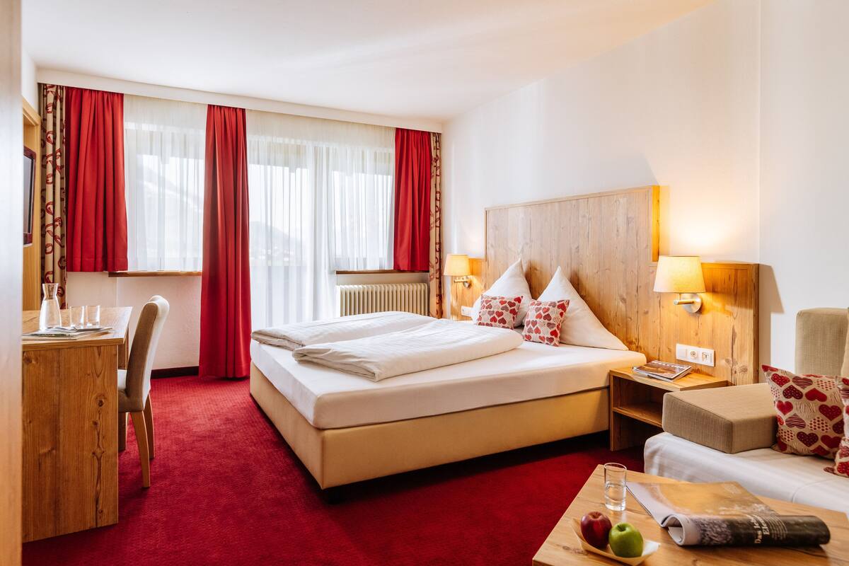 Quarto de hotel com uma cama grande, uma escrivaninha de madeira, uma cadeira, uma mesinha com uma cesta de frutas e um sofá.