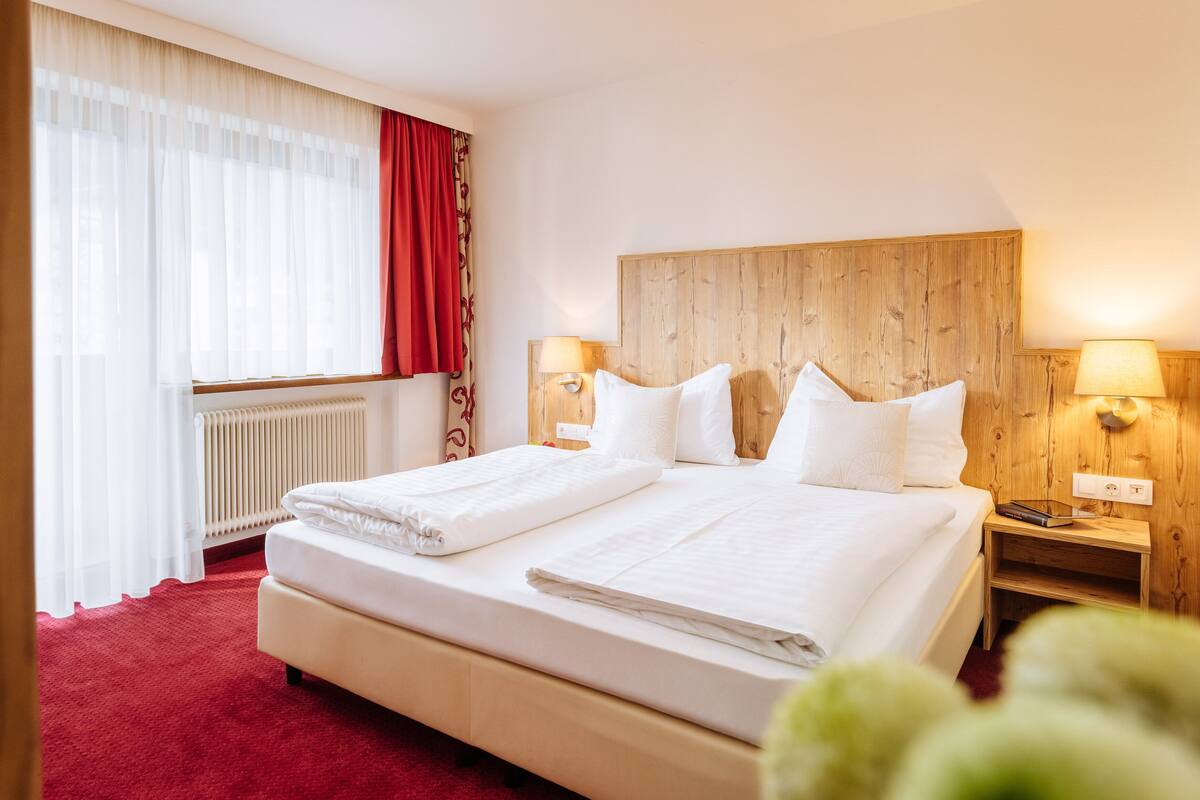 Quarto de hotel com uma cama grande, duas mesas de cabeceira com luminárias, uma cabeceira de madeira e uma janela com cortinas finas e um tecido vermelho.