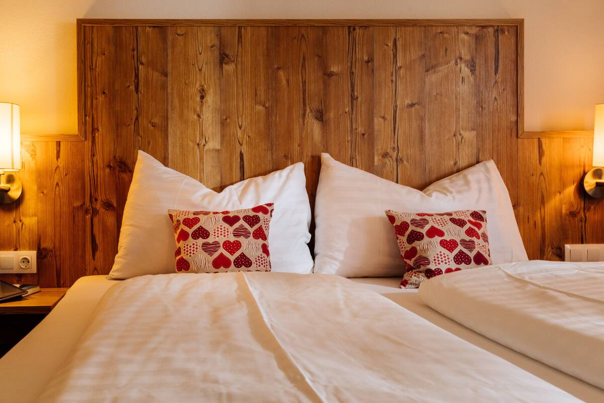Quarto de hotel com cabeceira de madeira, roupa de cama branca e almofadas estampadas com corações.