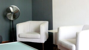 Quarto moderno com duas poltronas brancas, uma mesinha e um ventilador de chão em frente a uma parede cinzenta.