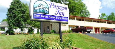 Placa do Paragon Inn com ilustração de montanha e árvores, rodeada por vegetação e um edifício ao fundo.