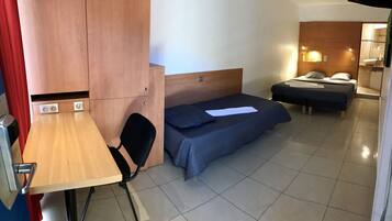 Quarto de hotel compacto com escrivaninha de madeira, uma cama de solteiro, uma cadeira e pequena área de cozinha.