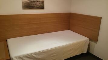 Quarto pequeno com uma cama de solteiro, cabeceira de madeira, colchão branco e uma fotografia emoldurada de um veleiro na parede.