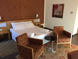 Quarto de hotel com cama, duas poltronas, uma mesinha, mesa de cabeceira, luminária e um quadro na parede.