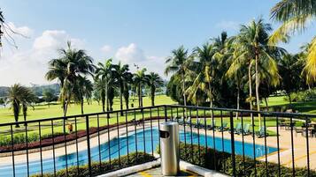 Área de piscina com uma lixeira de metal, cercada por uma grade, palmeiras e vista para um campo de golfe.