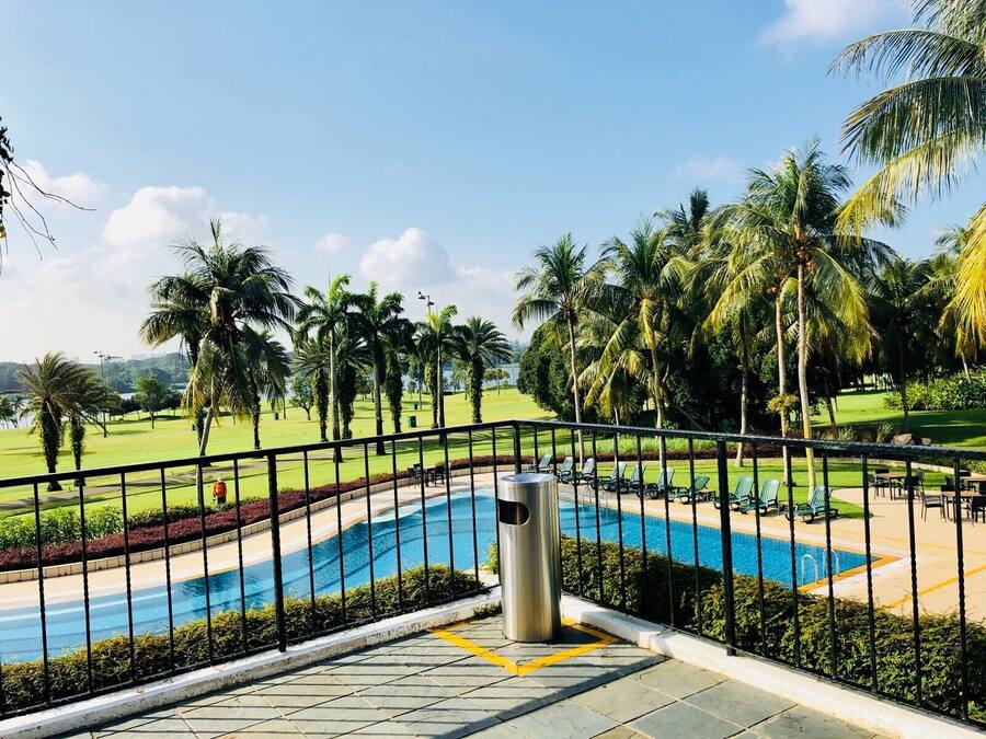 Área de piscina com uma lixeira de metal, cercada por uma grade, palmeiras e vista para um campo de golfe.